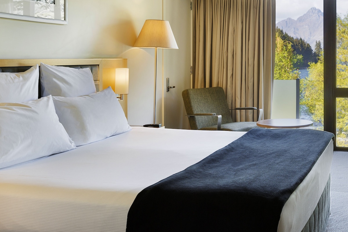 Quarto de hotel com cama, poltrona, luminária e vista para montanhas.