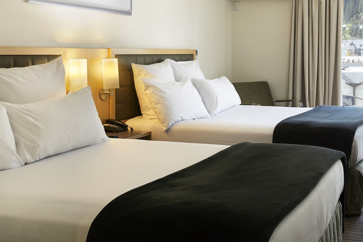 Quarto de hotel com duas camas, mesa de cabeceira, luminária e vista para o exterior.