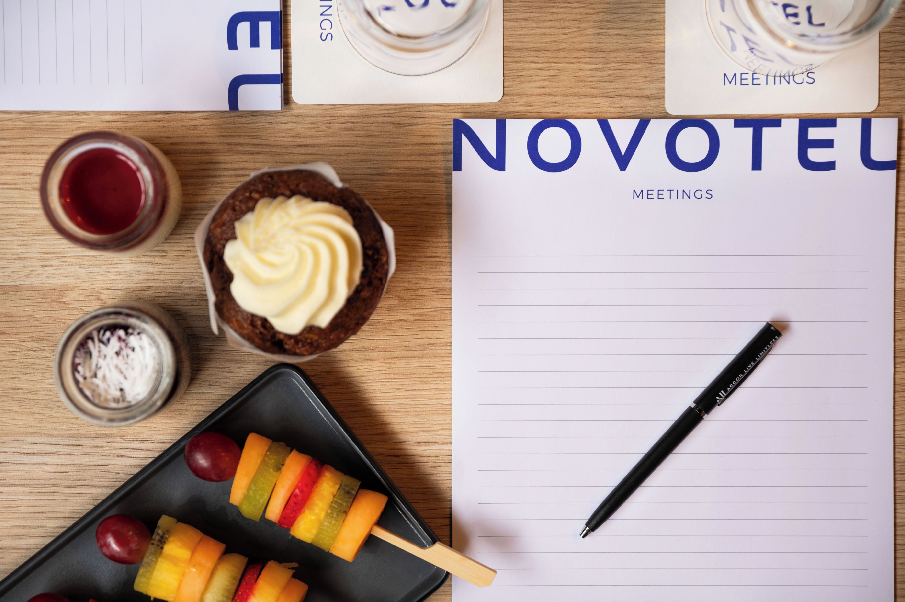 Uma mesa de madeira com um bloco de notas rotulado “NOVOTEL MEETINGS”, uma caneta preta, um copo com bebida vermelha, uma sobremesa com chantilly e um prato de espetinhos de fruta.