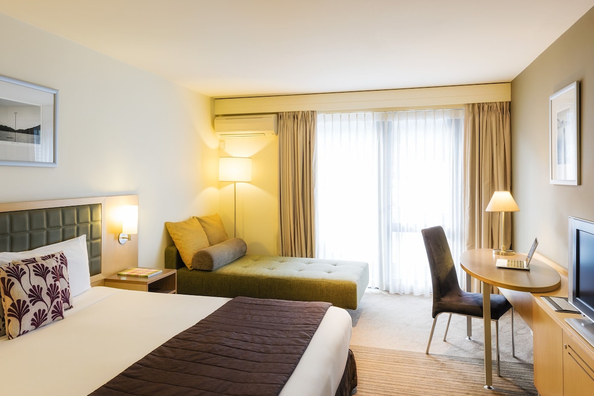 Quarto de hotel com cama, escrivaninha, laptop, cadeira, abajur e televisão.