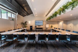 Uma sala de conferência com uma mesa comprida, cadeiras pretas, uma tela de TV e um letreiro com a inscrição 'NOVOTEL'.