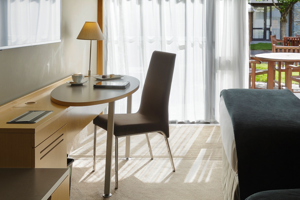 Quarto de hotel com cama, escrivaninha com luminária, cadeira e vista para o exterior através de cortinas translúcidas.