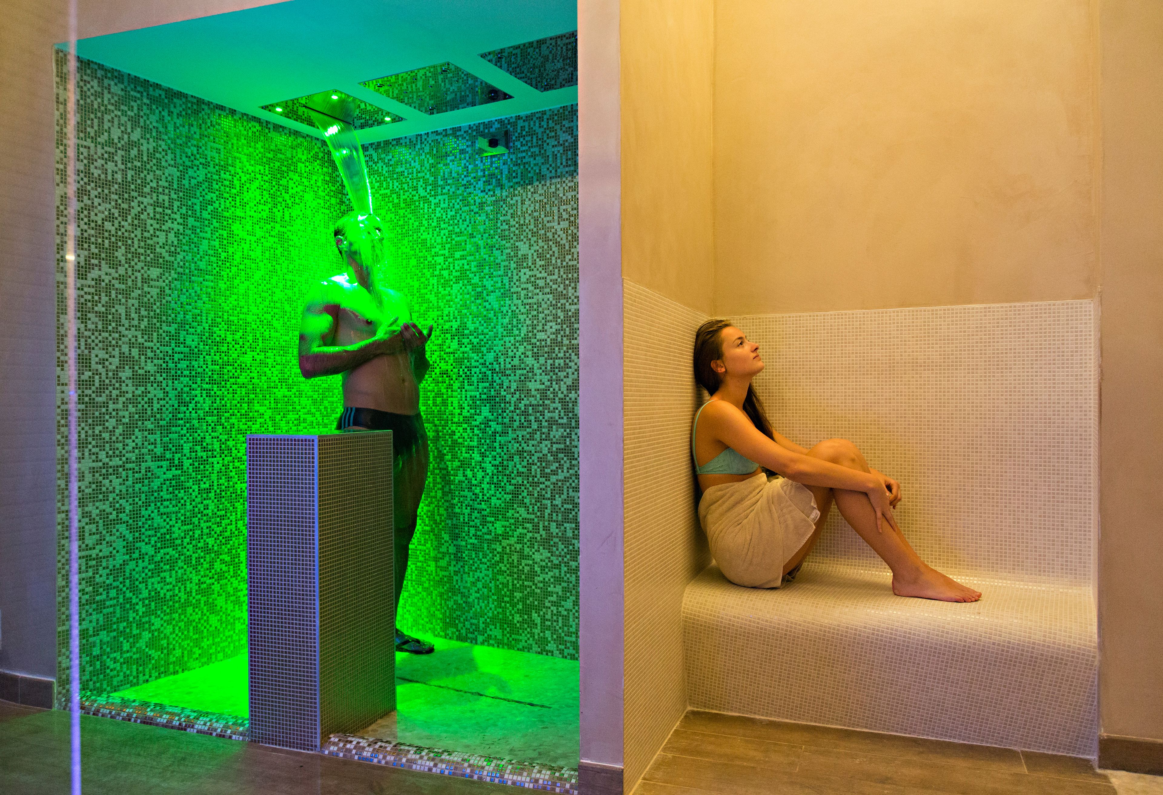 Casa de banho com área de duche iluminada em verde e zona de estar com parede revestida a azulejos.