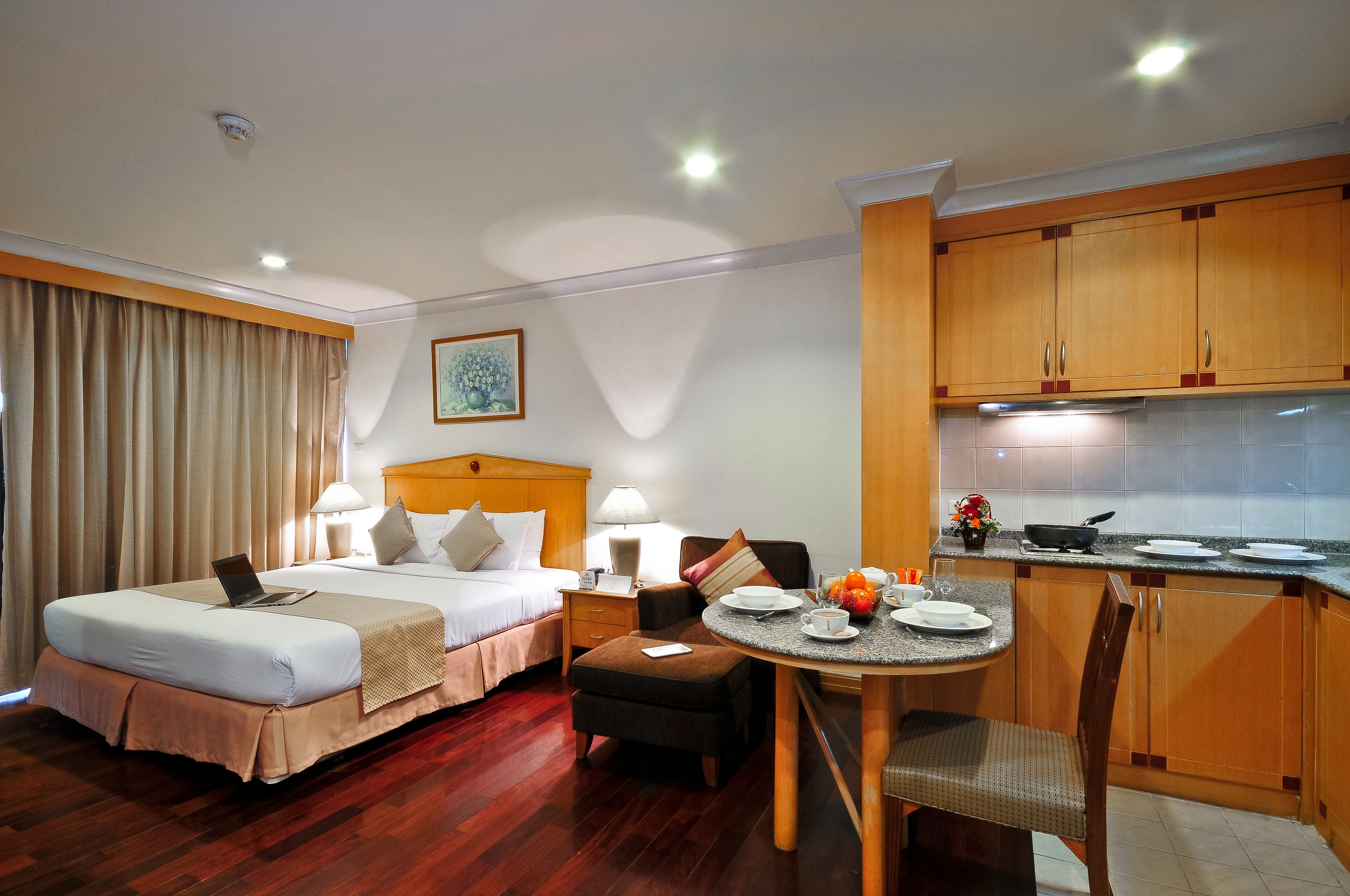 Quarto de hotel com cama, escrivaninha com laptop, mesa de jantar com pratos e uma saladeira, e cozinha com armários e fogão.