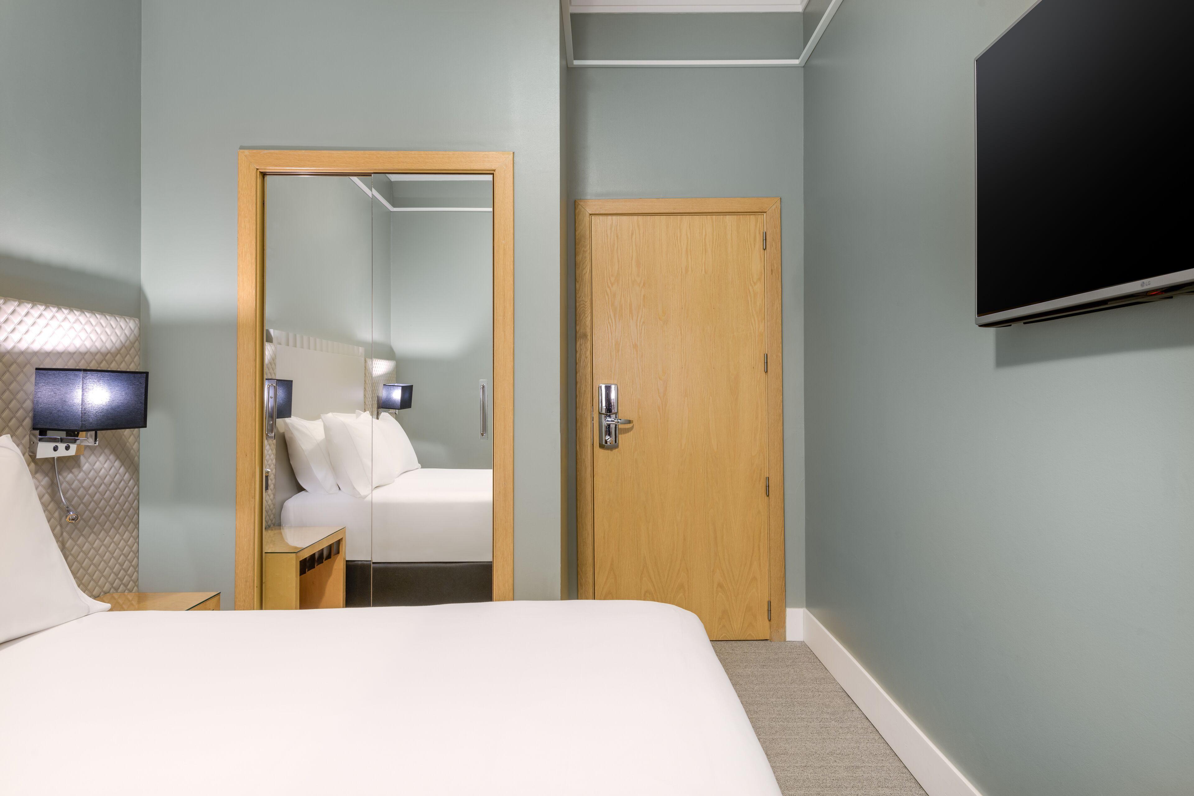 Quarto de hotel com uma cama, porta de madeira, televisão na parede e mesa de cabeceira com abajur.