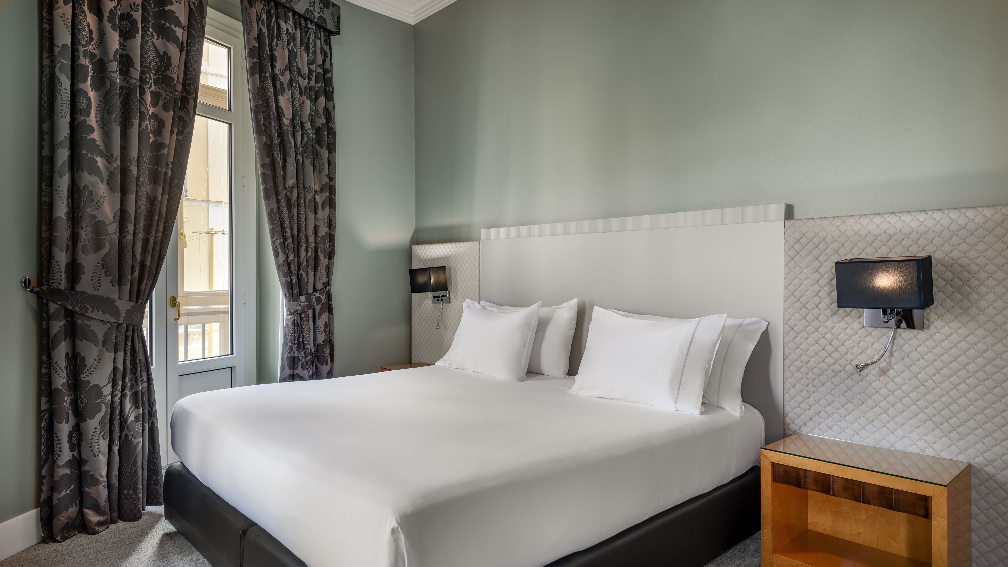Cama bem arrumada com lençóis brancos, mesa de cabeceira de madeira com tampo de vidro e luminária fixada na parede.