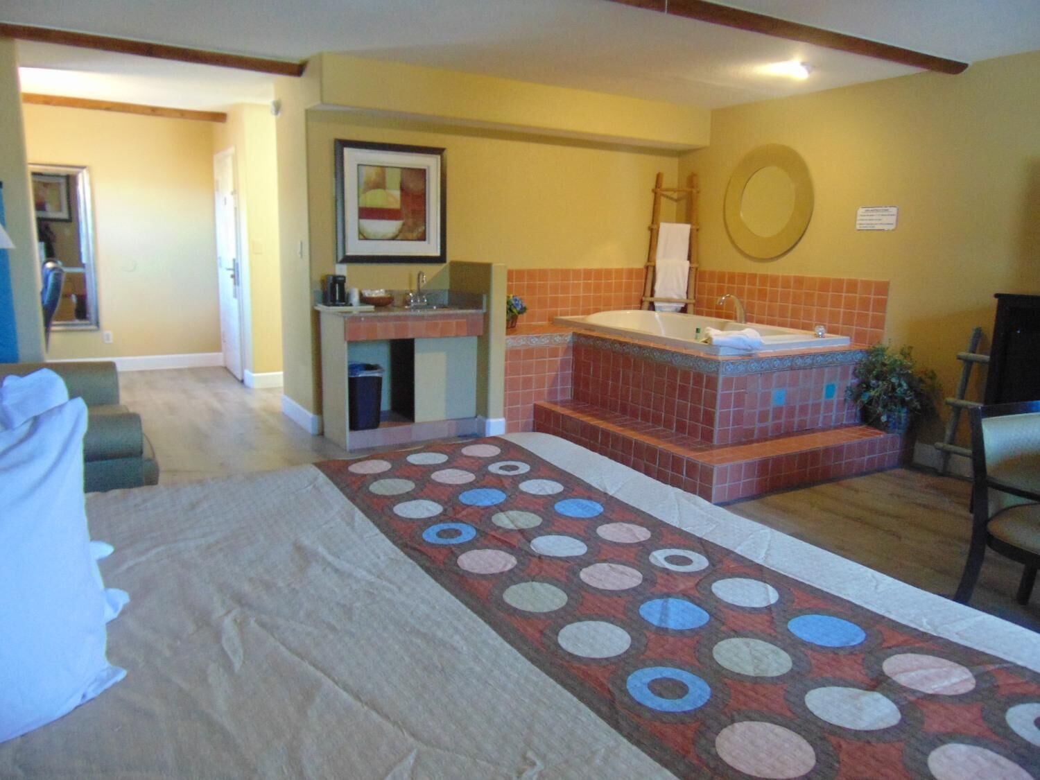 Quarto de hotel com cama, pequeno espaço de cozinha, banheiro com banheira e tapete colorido.
