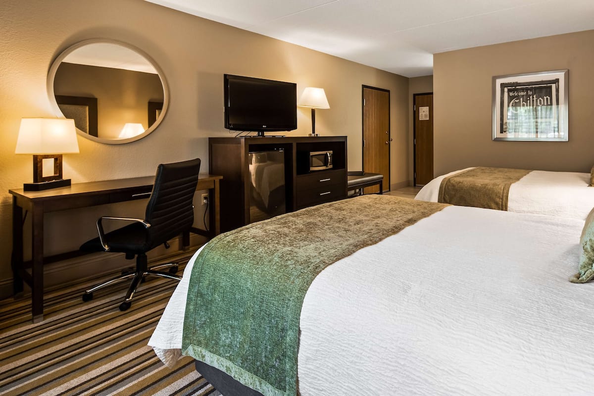 Quarto de hotel com duas camas, uma escrivaninha com computador e televisão, micro-ondas e um espelho redondo.