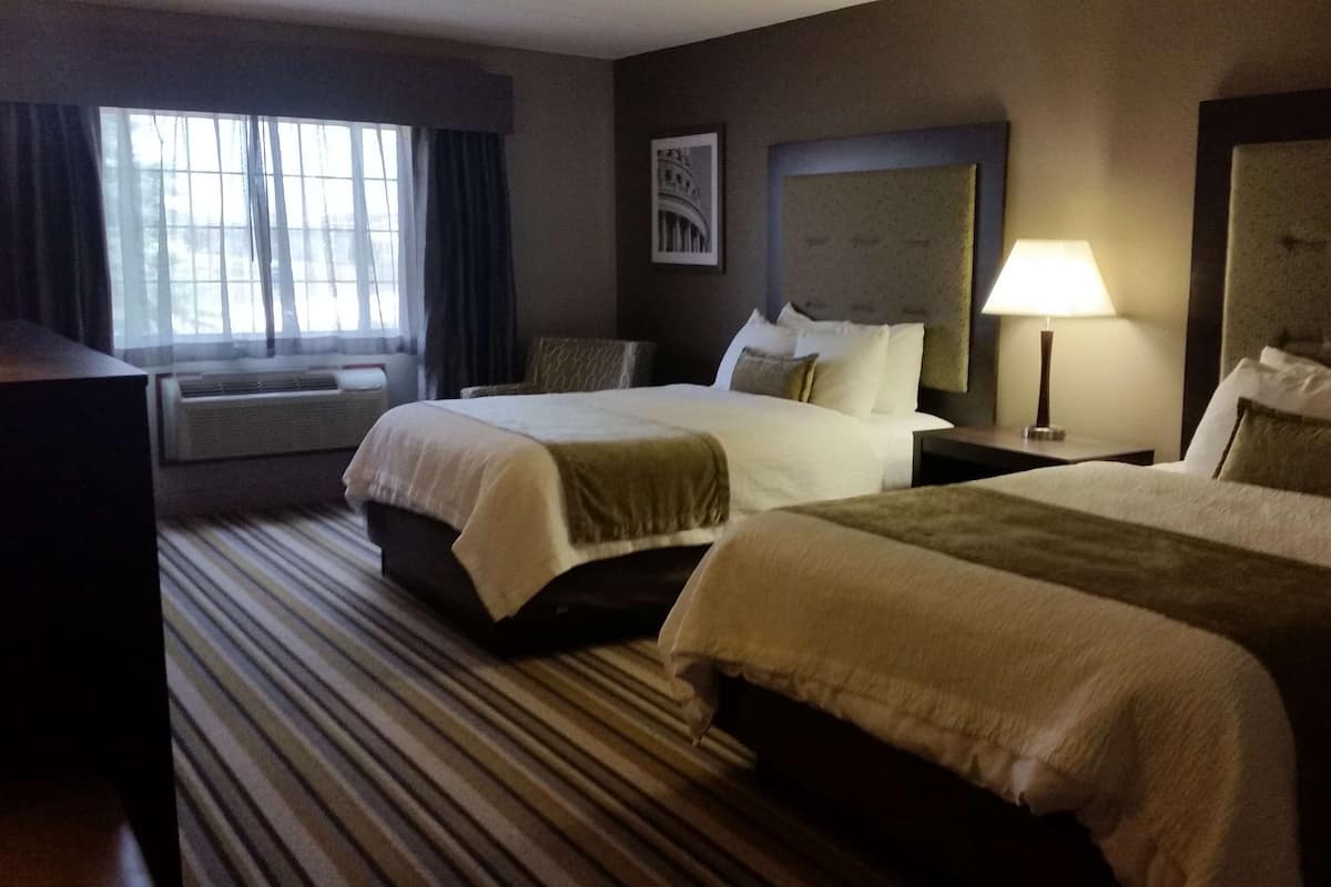 Quarto de hotel com duas camas, uma escrivaninha, uma luminária e uma janela com cortinas.