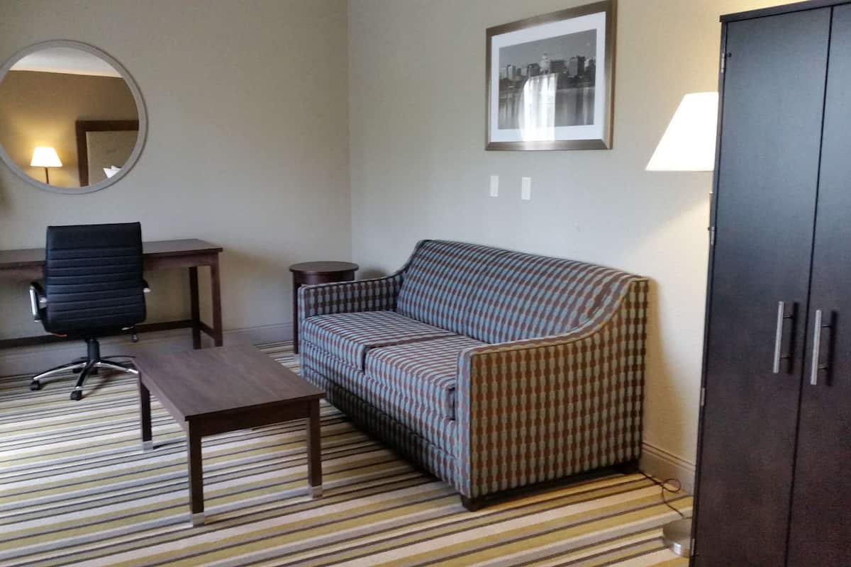 Quarto de hotel com carpete listrado, um sofá, uma escrivaninha com cadeira, um espelho redondo, uma luminária e um guarda-roupa.