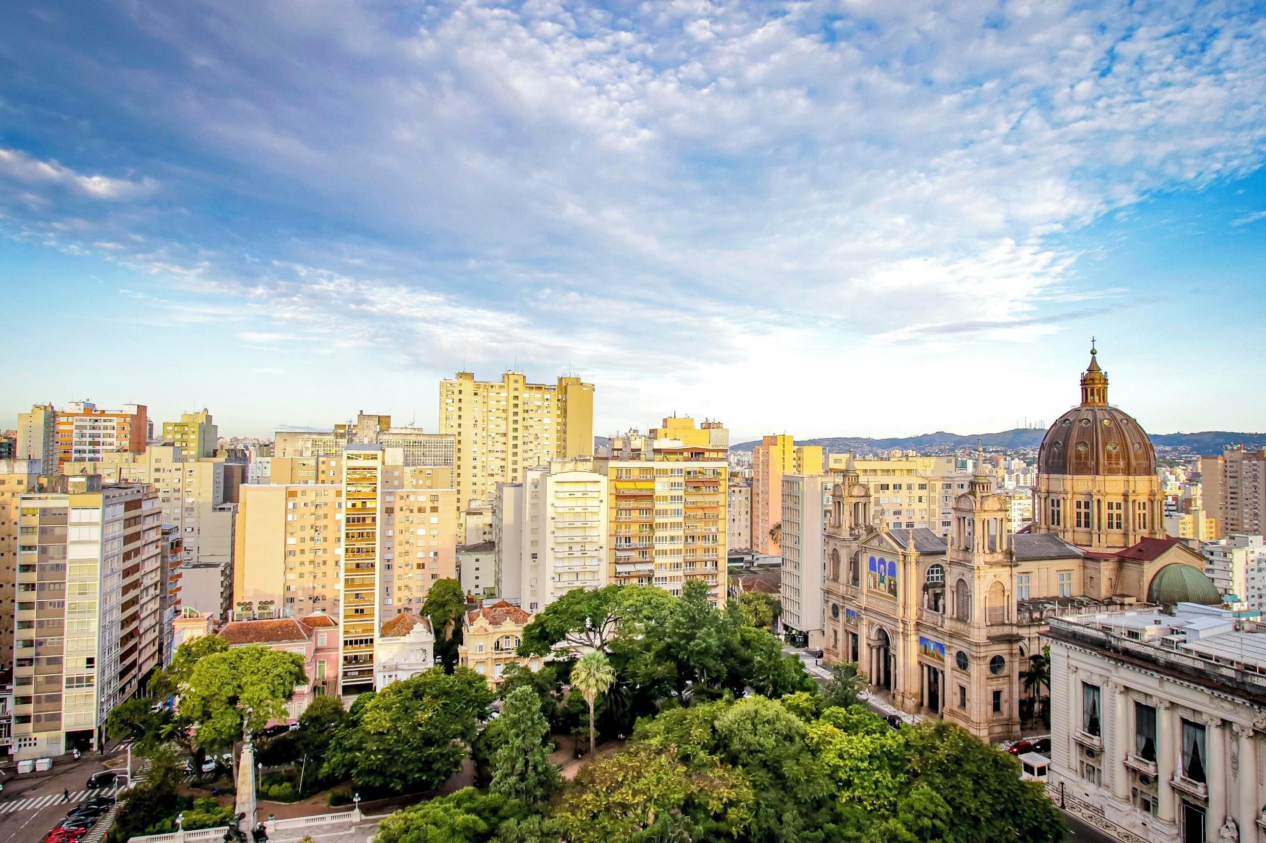 Paisagem urbana com edifícios altos, um edifício histórico imponente com cúpula e árvores verdes exuberantes em primeiro plano.