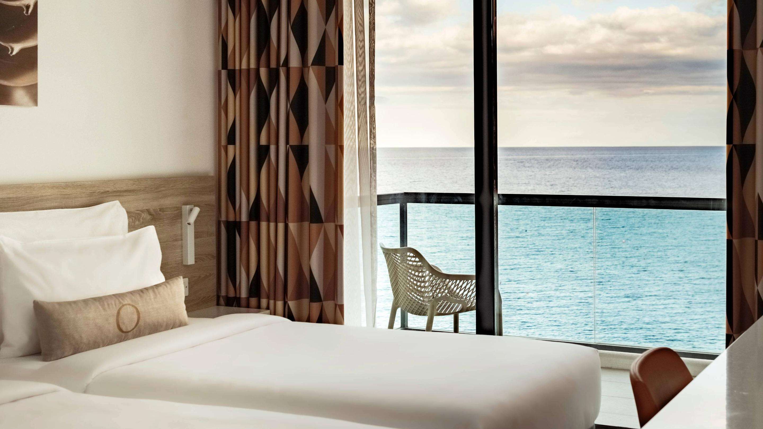 Quarto de hotel com uma cama grande, vista para o oceano e varanda com uma cadeira.