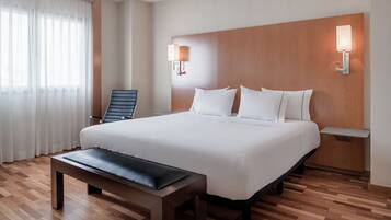 Quarto de hotel com uma cama grande, um banco, duas luminárias fixadas na parede e uma cabeceira de madeira.