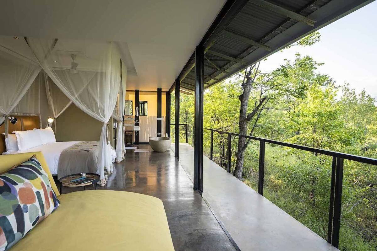 Quarto com cama, mesa de cabeceira e uma grande janela com vista para uma floresta.