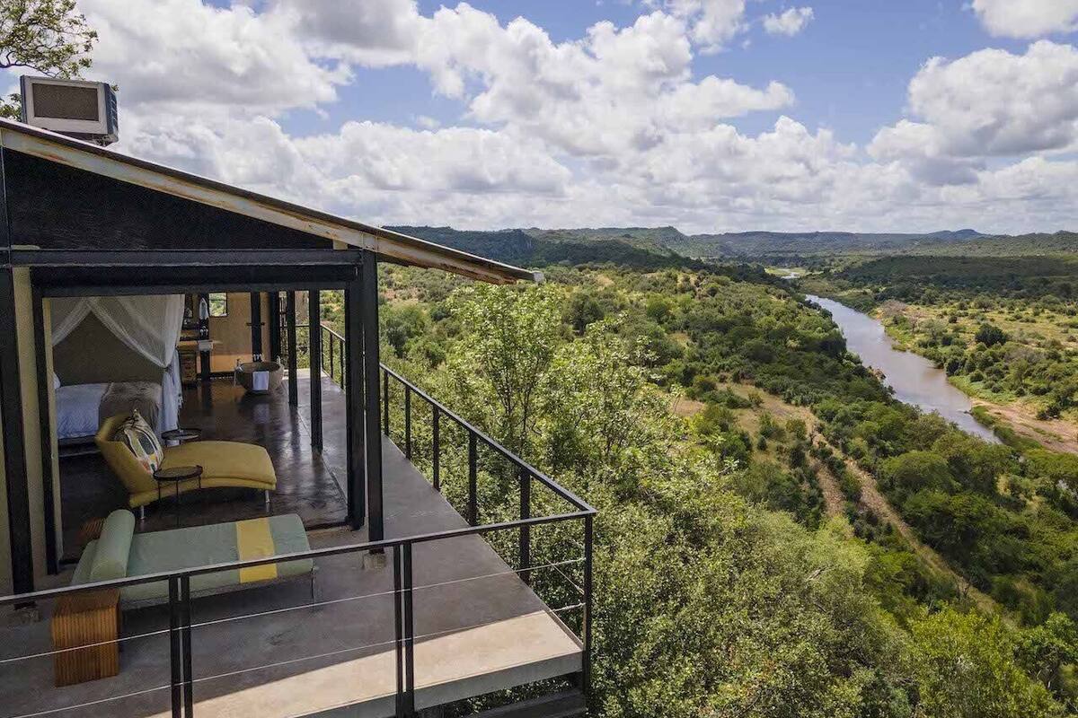 Uma área de estar moderna ao ar livre, com sofá e cama, com vista para um rio e paisagem montanhosa.