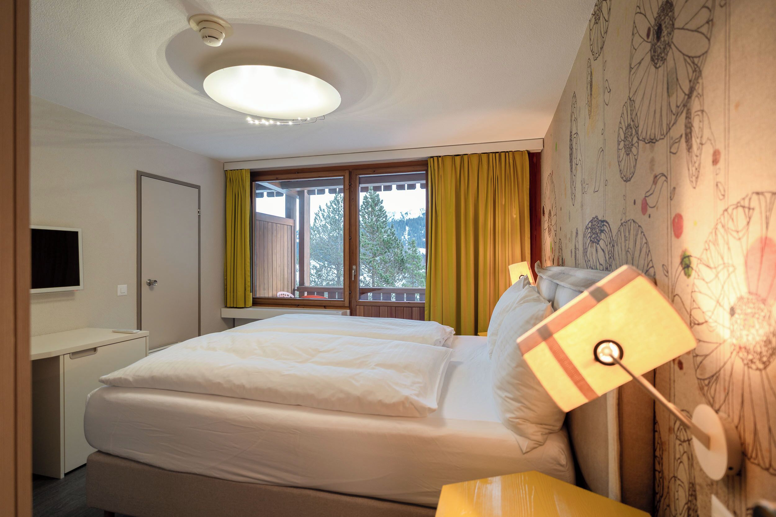 Quarto com cama, escrivaninha, janela com cortinas e parede estampada com flores.