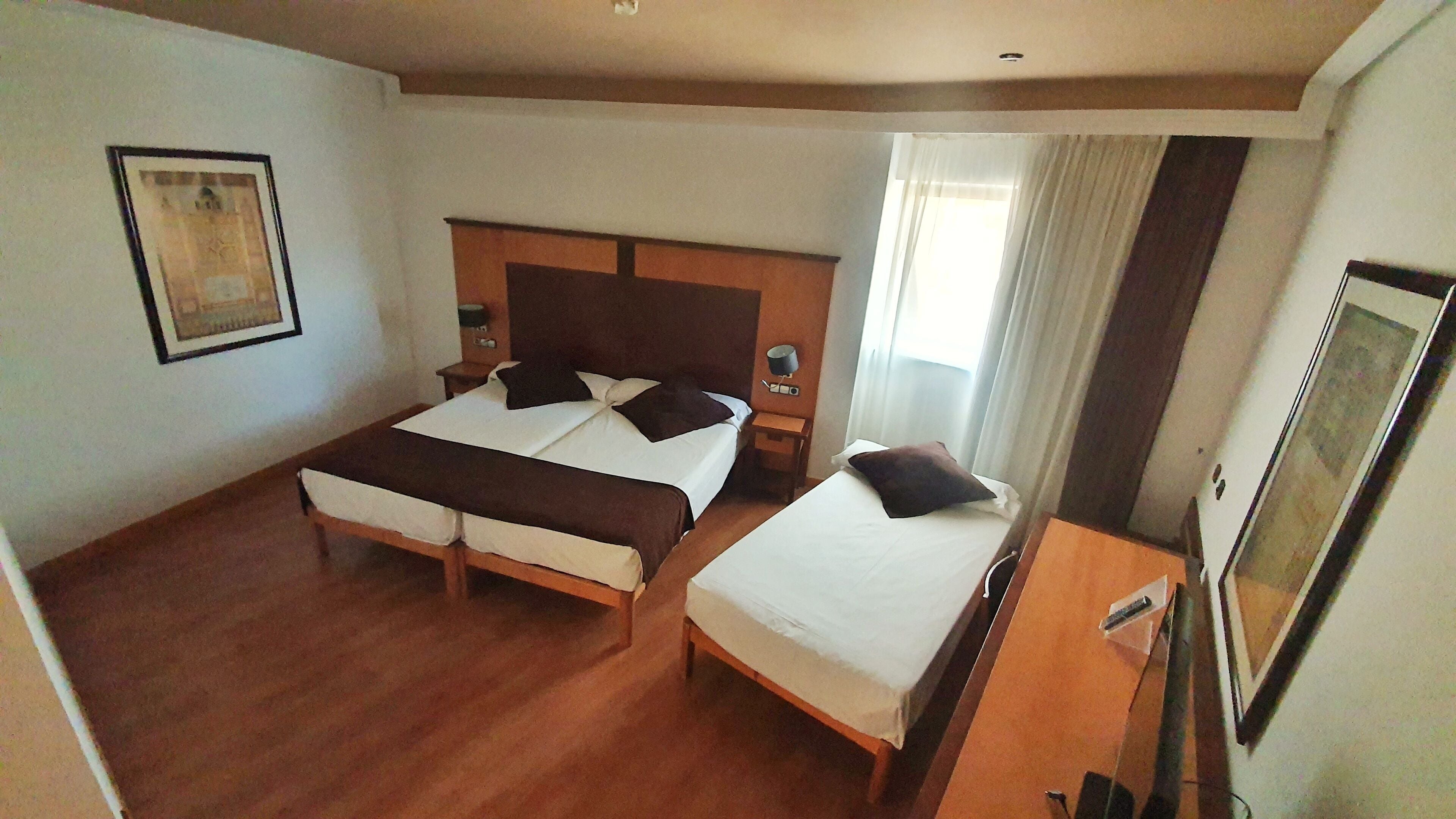 Quarto de hotel com duas camas, cabeceiras de madeira, uma mesa de cabeceira com abajur e um quadro na parede.