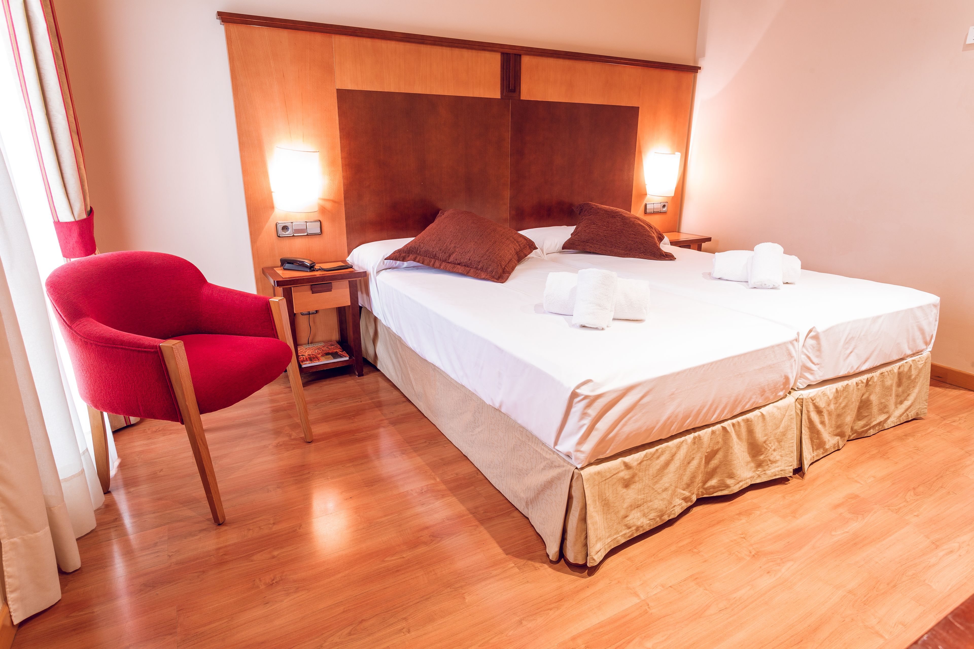 Quarto de hotel com cabeceira de madeira, uma cadeira vermelha, cama com lençóis brancos e almofadas castanhas, e duas luminárias de cabeceira.