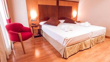 Quarto de hotel com cabeceira de madeira, uma cadeira vermelha, cama com lençóis brancos e almofadas castanhas, e duas luminárias de cabeceira.