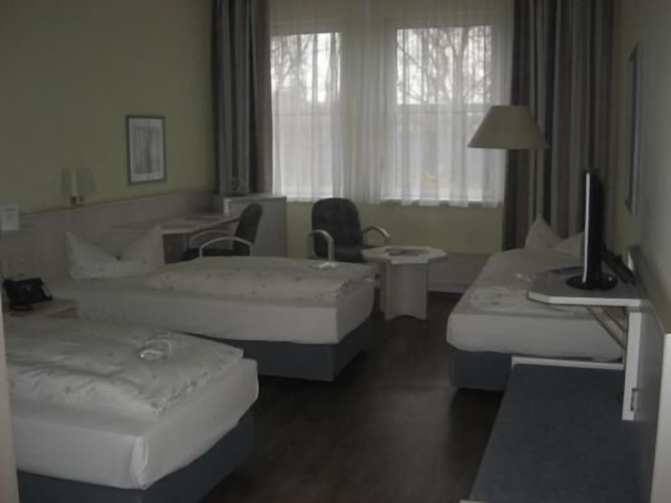 Quarto de hotel com duas camas, uma mesa, uma cadeira, uma luminária e uma janela com cortinas.