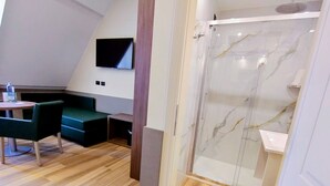 Quarto de hotel moderno com piso de madeira, um sofá verde, uma mesinha, televisão fixada na parede e box de vidro.