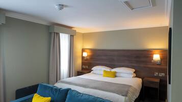 Quarto de hotel com uma cama grande, duas mesas de cabeceira, um criado-mudo, uma janela com cortinas e uma luminária no teto.