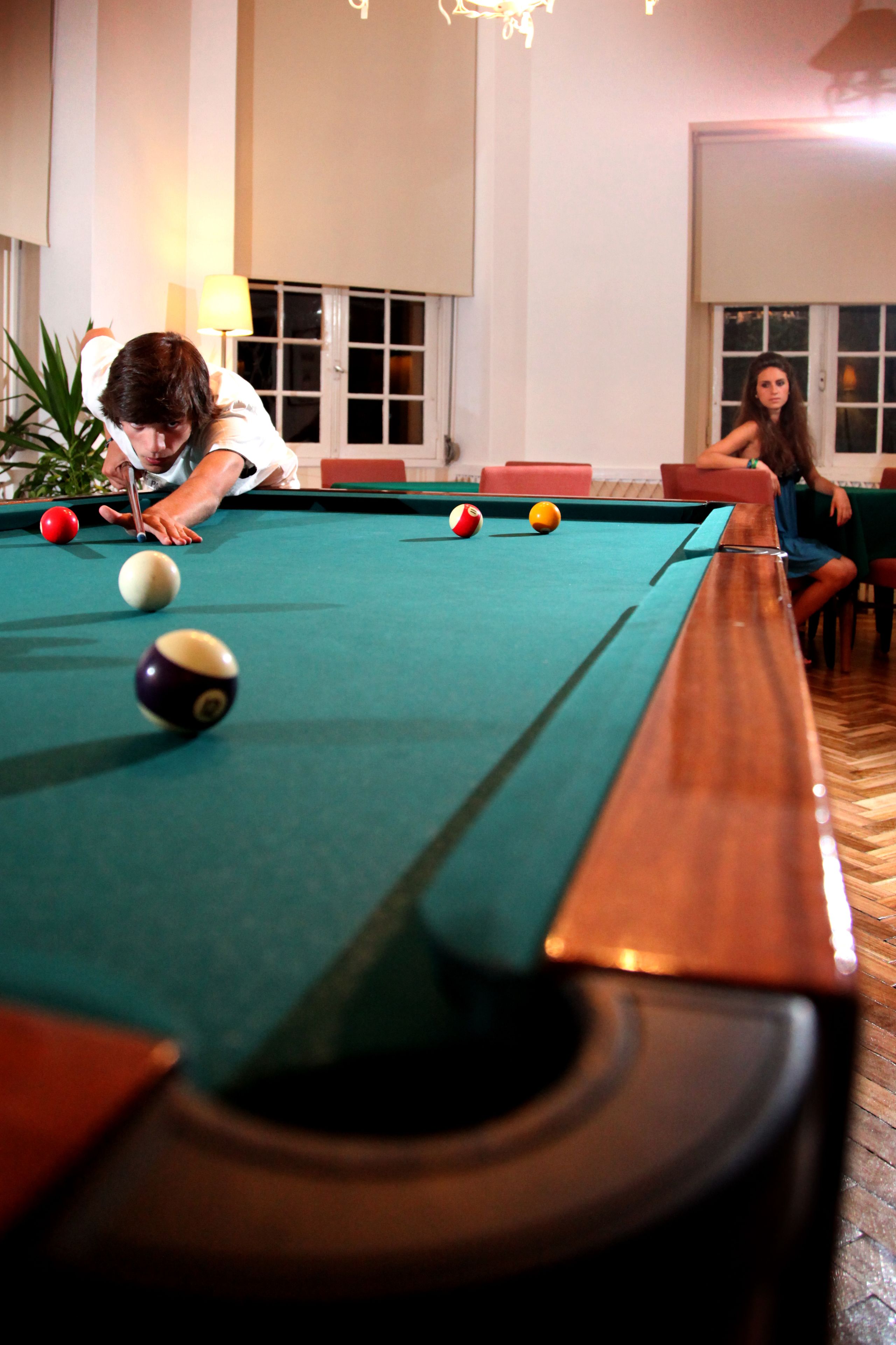 Uma mesa de bilhar em um ambiente com piso de madeira e uma luminária ao fundo.