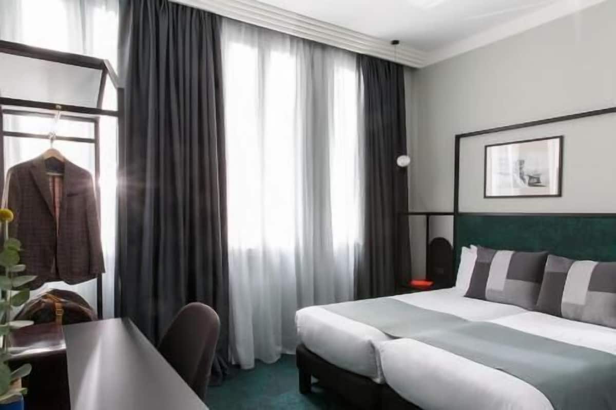 Quarto de hotel com cama, escrivaninha, cadeira, porta-roupas e janela com cortinas.