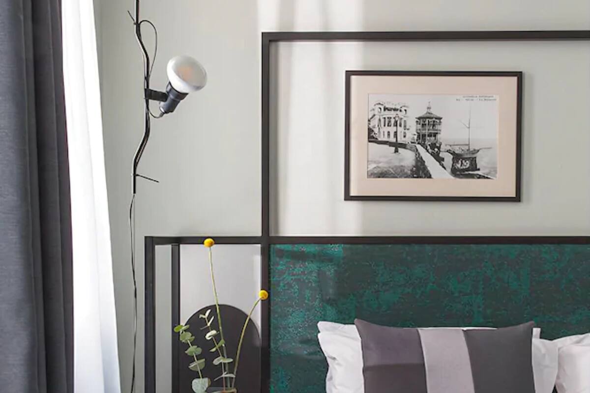 Quarto de hotel com cama, um quadro na parede, mesa de cabeceira com abajur e uma planta.