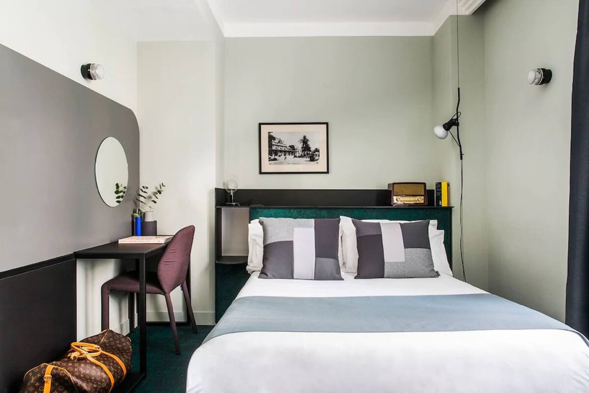Quarto de hotel com uma cama, uma escrivaninha, uma cadeira, um abajur e um quadro pendurado na parede.