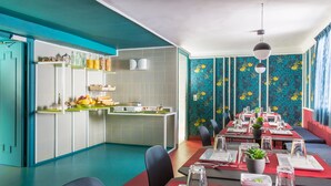 Uma sala de jantar com uma mesa comprida posta para refeição, área de cozinha com bancada e armários, e parede com padrão floral.