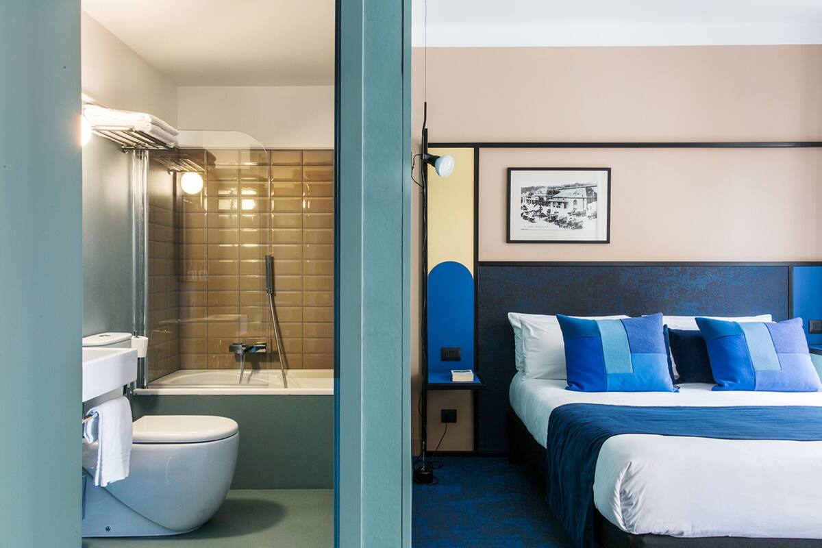 Quarto de hotel com banheiro, cama com roupa de cama azul e um quadro pendurado na parede.