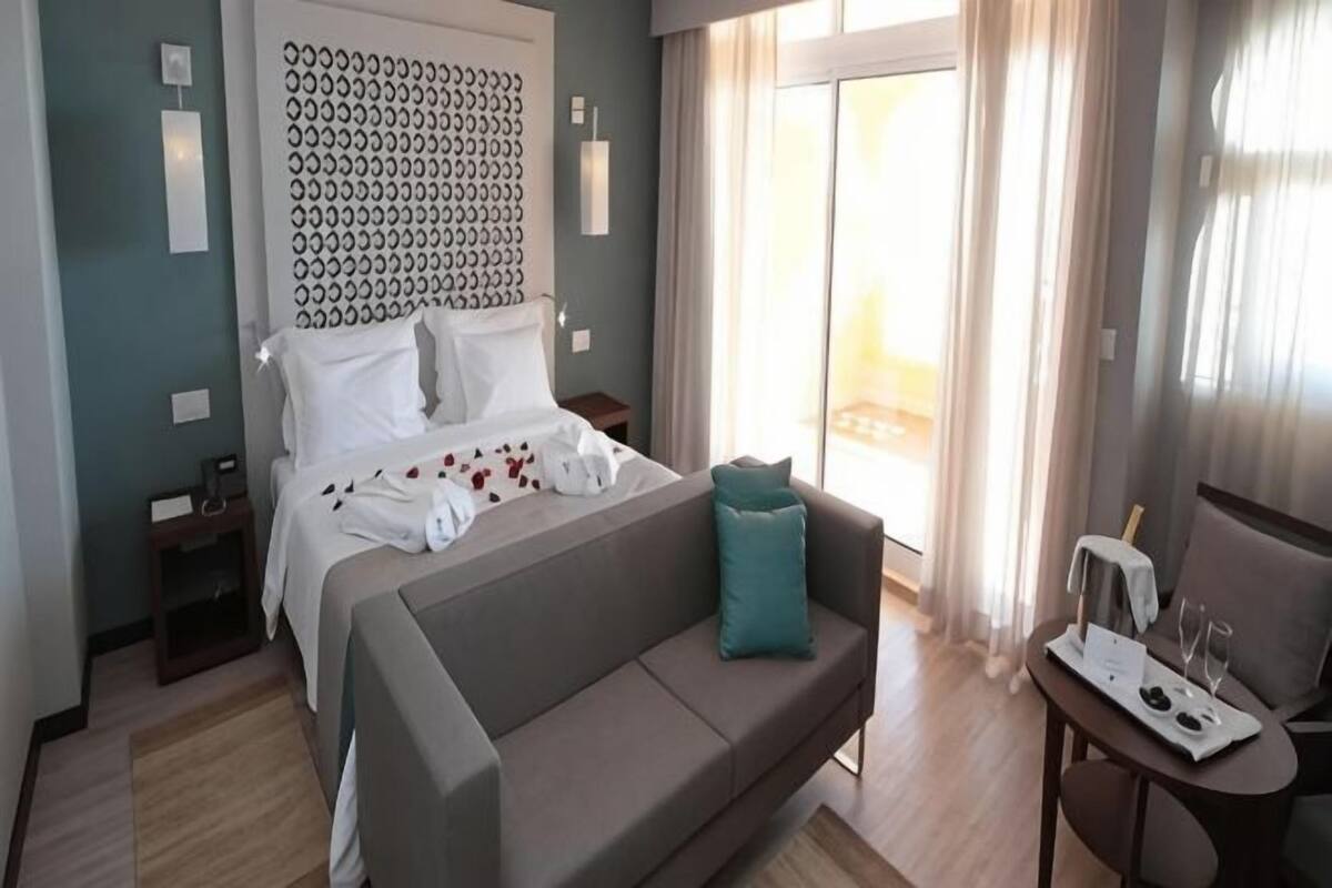 Quarto de hotel com cama, sofá, uma mesinha com copos e vista para o exterior.