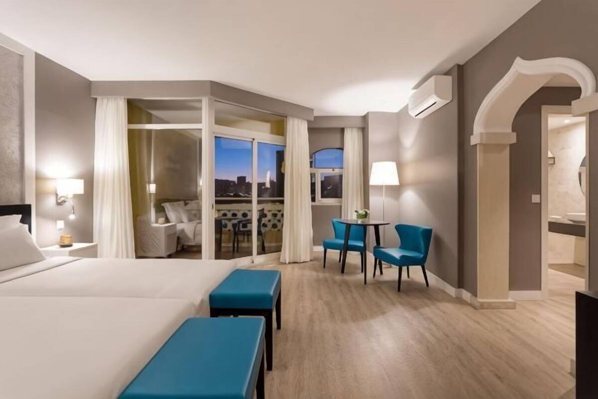 Quarto de hotel com cama, escrivaninha, cadeira, varanda e vista para a cidade, além de banheiro.