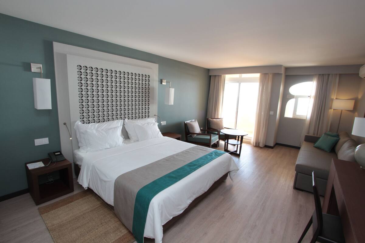 Quarto de hotel com uma cama grande, uma escrivaninha, uma cadeira, um sofá e uma janela com cortinas.