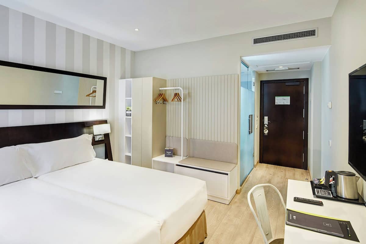 Quarto de hotel com uma cama, uma escrivaninha com cadeira, um espelho, um guarda-roupa e uma porta.