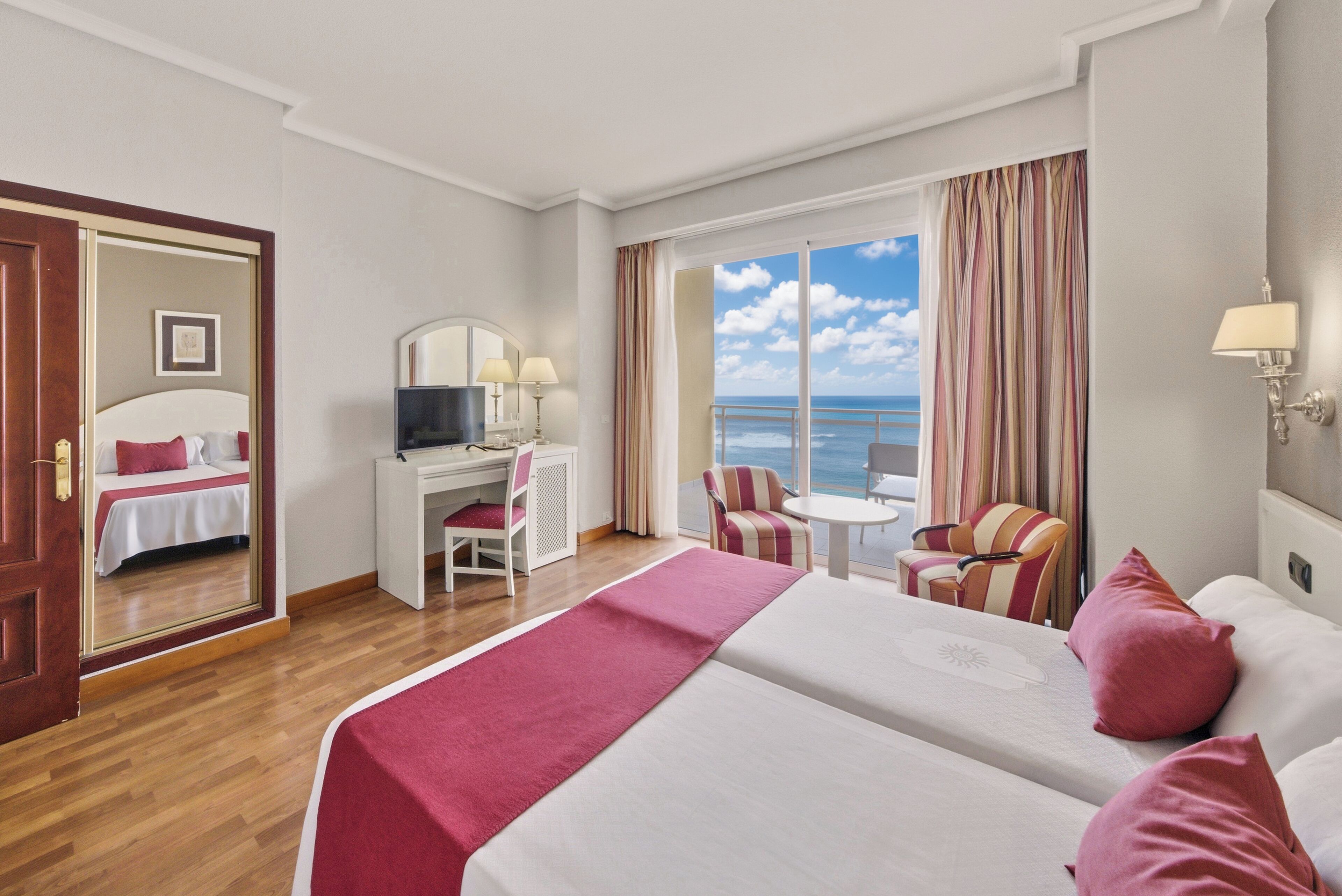 Quarto de hotel com duas camas, uma mesa de trabalho com televisão, uma cadeira e vista para o oceano.