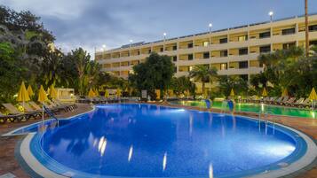Piscina com espreguiçadeiras e guarda-sóis, cercada por palmeiras e um prédio de hotel.