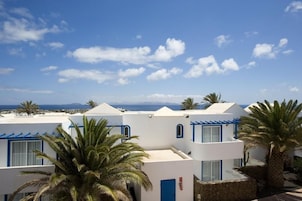 Hotel à beira-mar com edifícios brancos e detalhes em azul, apresentando palmeiras e vista para o mar e montanhas.
