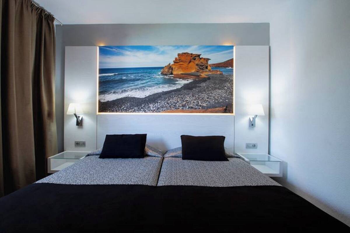 Quarto de hotel com uma cama, duas almofadas, uma mesa de cabeceira, uma luminária fixada na parede e um quadro grande com vista para a paisagem costeira.