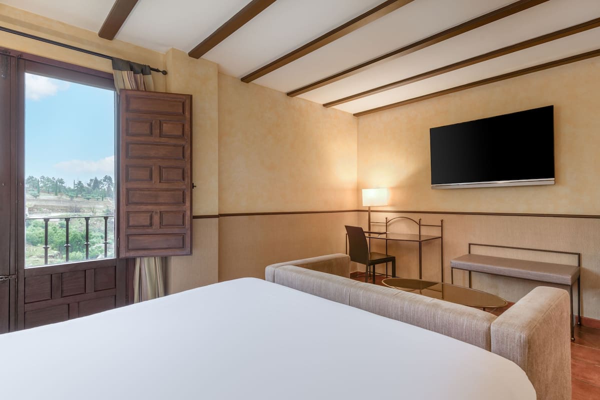 Quarto de hotel com cama, TV fixada na parede, escrivaninha com cadeira e janela com vista para o verde.