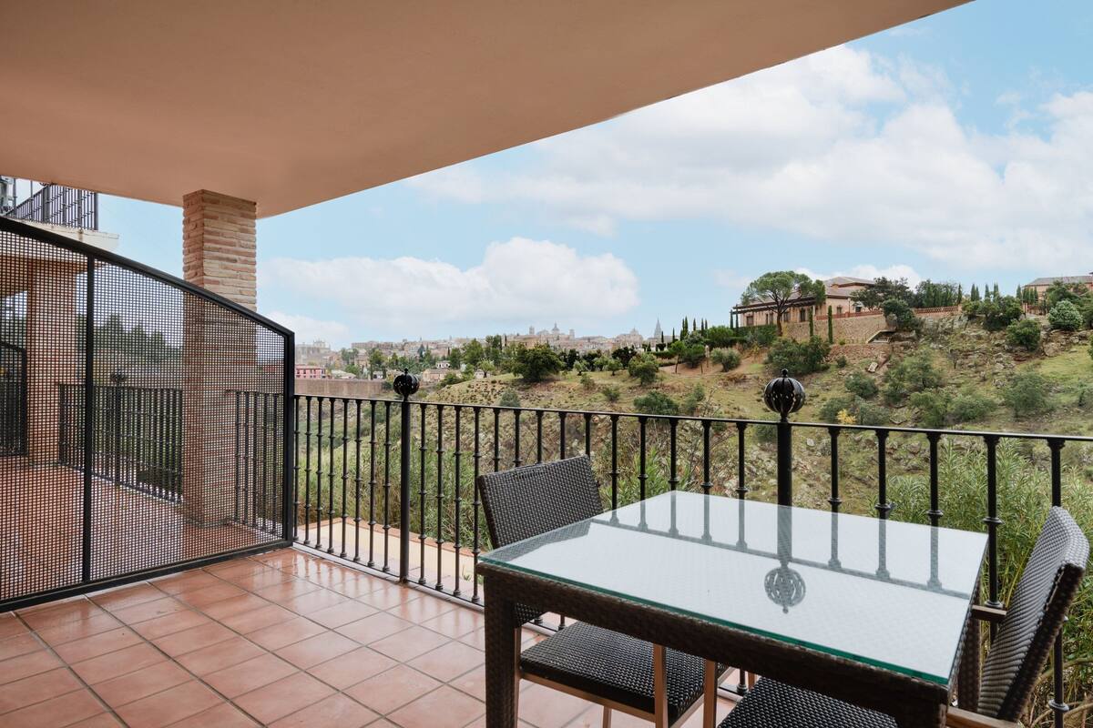 Varanda com mesa e cadeiras de vidro, guarda-corpo metálico e vista para a cidade e áreas verdes.
