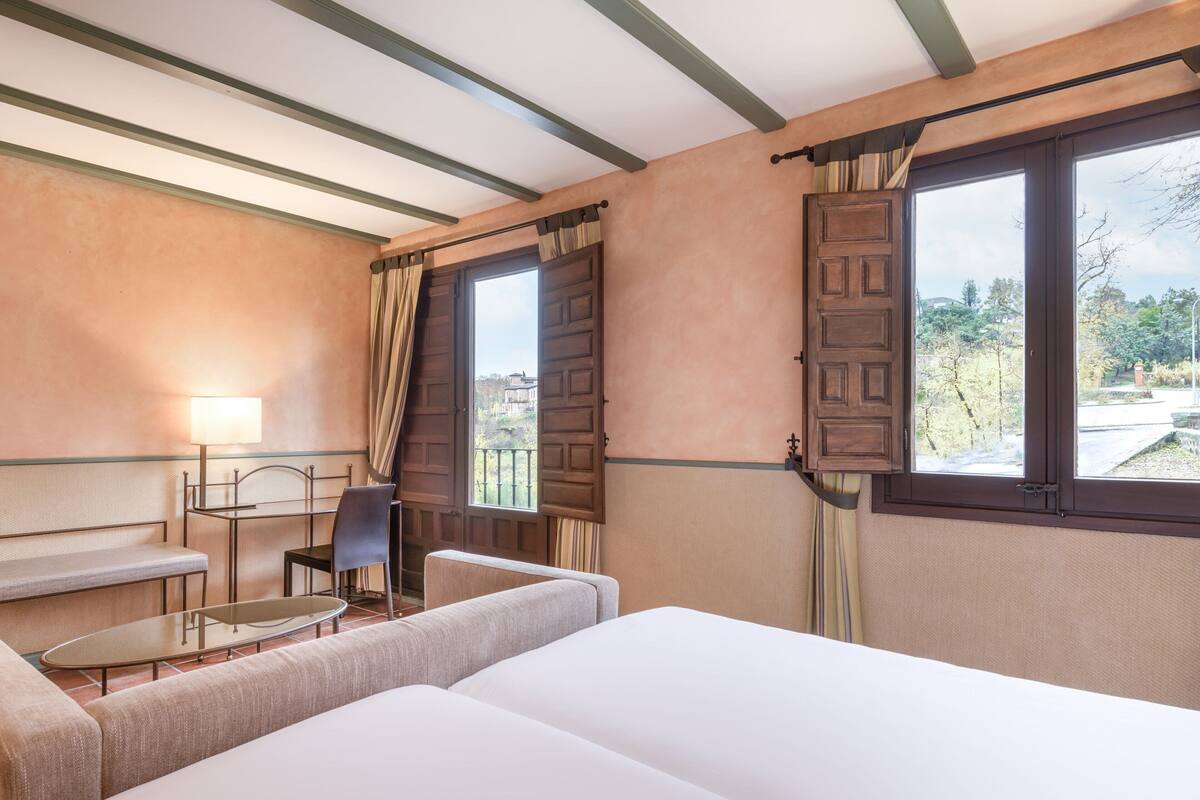 Quarto de hotel com cama, sofá, mesa pequena, cadeira e janela com cortinas.