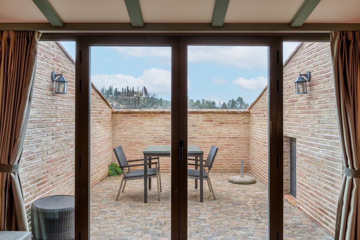 Vista através de uma porta de vidro para um pátio com parede de tijolos, uma mesa e cadeiras.