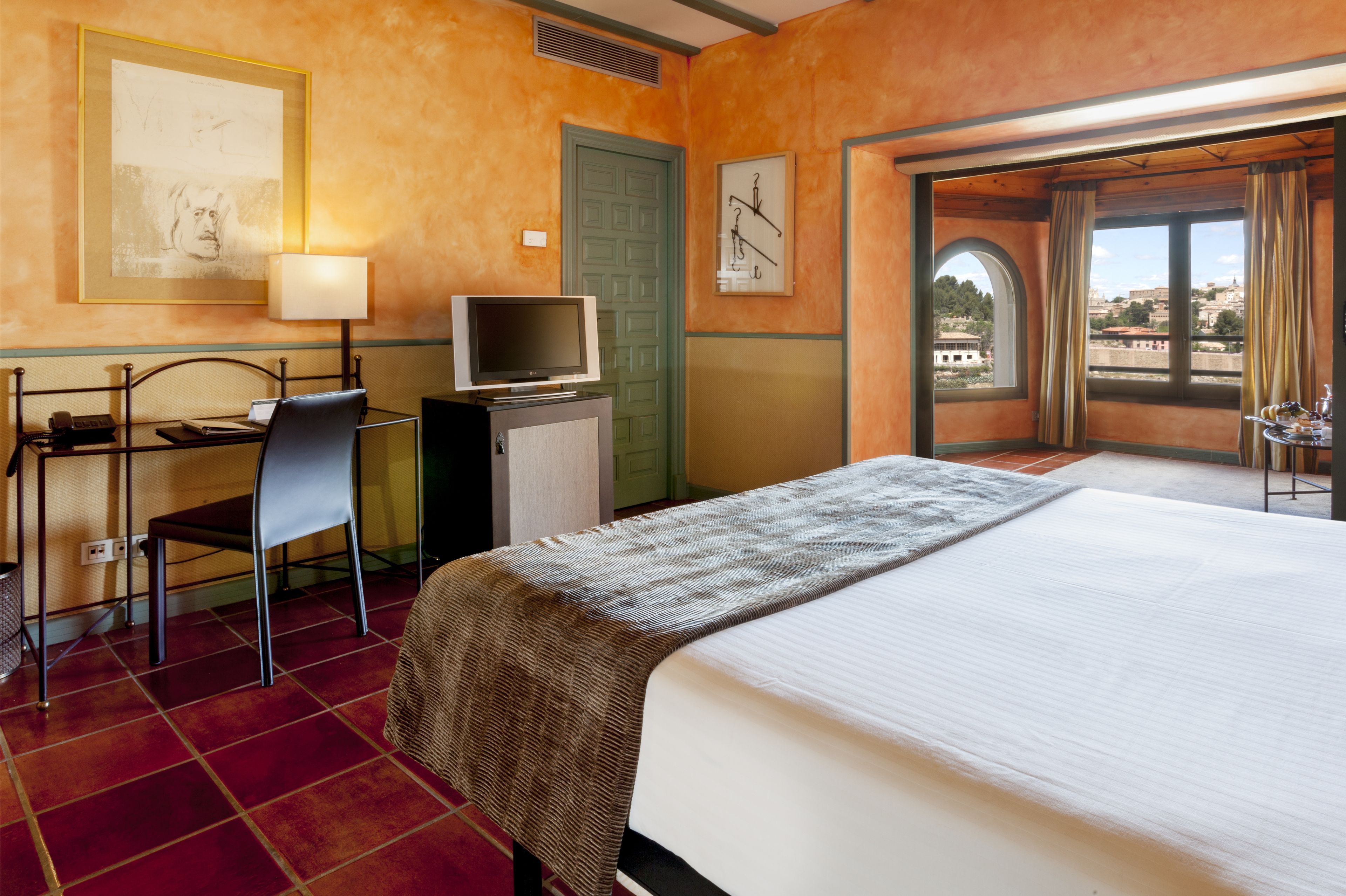 Quarto de hotel com cama, escrivaninha, cadeira, TV e vista para a cidade pela janela.