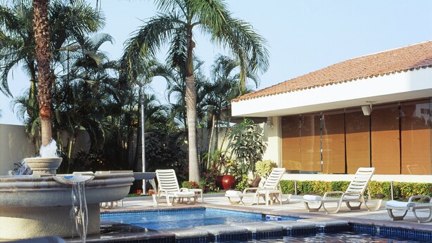 Uma área de piscina com espreguiçadeiras, uma fonte e um edifício com telhado em azulejos.