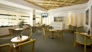 Um saguão de hotel com mesas e cadeiras redondas, um espaço de bar com garrafas e uma tela de televisão.