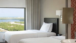 Quarto de hotel com duas camas, uma janela grande com vista para o mar e vegetação, banheiro com pia e espelho, e uma luminária na parede.