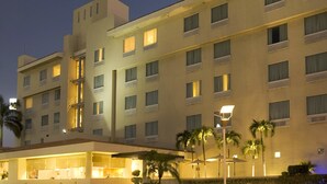 Um hotel de vários andares com fachada plana, várias janelas e uma área de entrada bem iluminada, com palmeiras e iluminação externa.