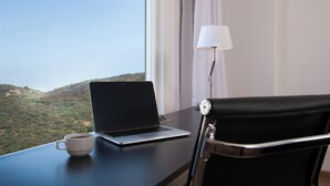 Um laptop sobre uma mesa com uma xícara, uma luminária e uma cadeira, com vista para uma paisagem acidentada.
